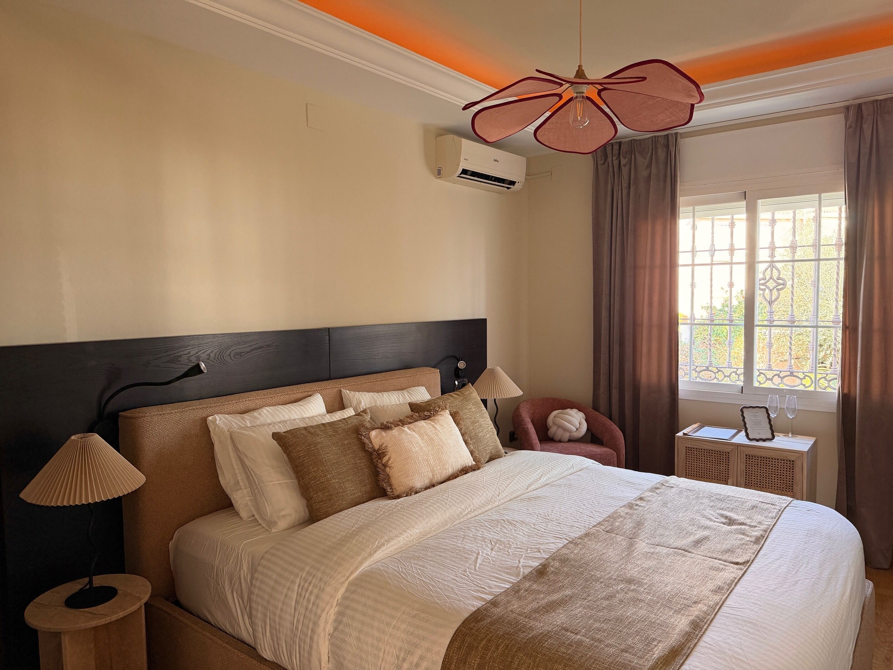 Quarto com uma cama grande, abajur, luminária decorativa no teto, janela com cortinas e aparelho de ar condicionado.