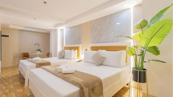 Quarto de hotel com duas camas, uma mesinha com uma planta e um vaso grande com planta junto à janela.