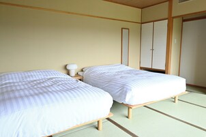 Quarto em estilo japonês tradicional com tatames, duas camas de solteiro com roupa de cama branca, uma mesa de cabeceira de madeira e um guarda-roupa com portas de correr.
