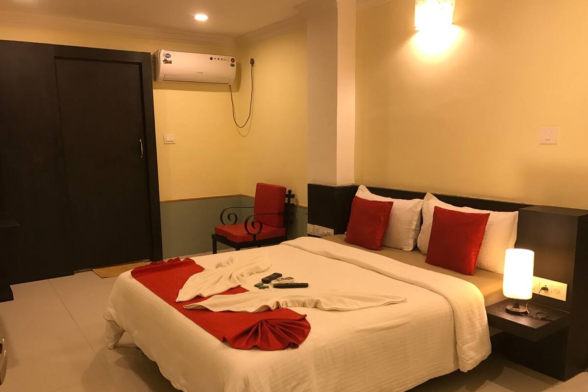 Quarto de hotel com uma cama grande, uma mesa de cabeceira com abajur, uma cadeira, um aparelho de ar condicionado e uma porta.