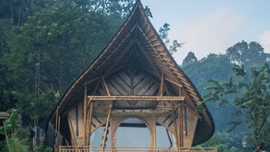 Estrutura tradicional de bambu com telhado de palha, rodeada por vegetação exuberante e um pequeno lago.