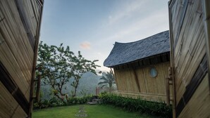 Uma casa tradicional de bambu com telhado de palha, um caminho de pedra que a conduz e vegetação exuberante ao redor da propriedade.