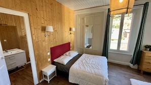 Quarto com parede de madeira, cama com roupa de cama branca, mesa de cabeceira, penteadeira, espelho e janela com cortinas.