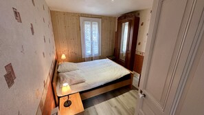 Quarto com cama de madeira, duas luminárias de cabeceira, janela com cortinas de renda e porta com espelho.