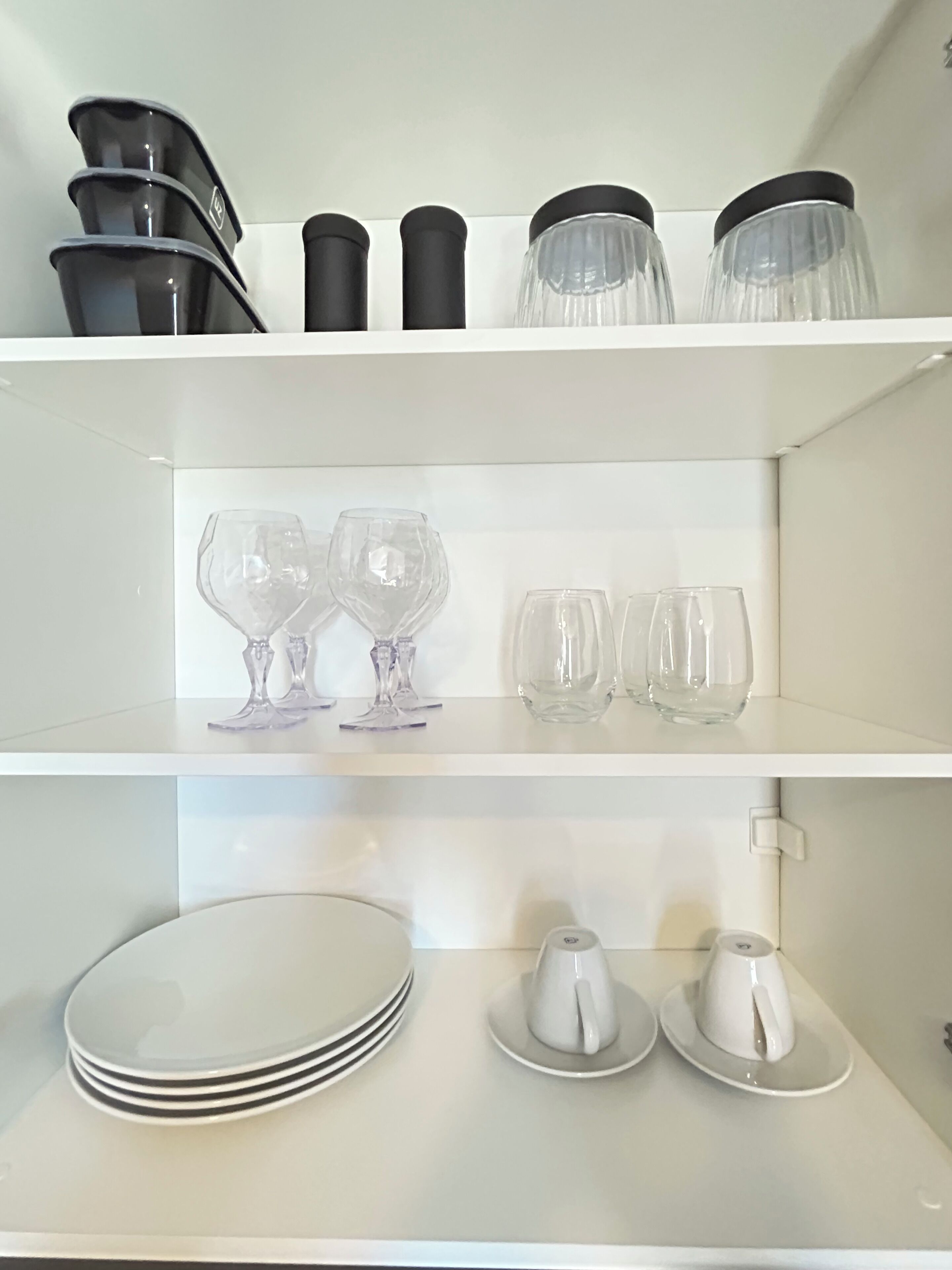 Uma cozinha bem organizada com prateleiras brancas que exibem utensílios de cozinha, incluindo recipientes pretos e transparentes, copos de vinho e pratos brancos.