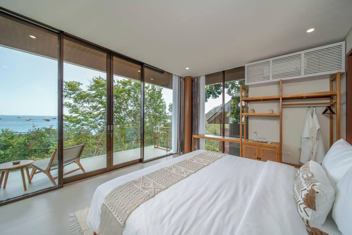 Quarto com uma cama grande, mesa de cabeceira de madeira, guarda-roupa e vista para o mar através de janelas do chão ao teto.