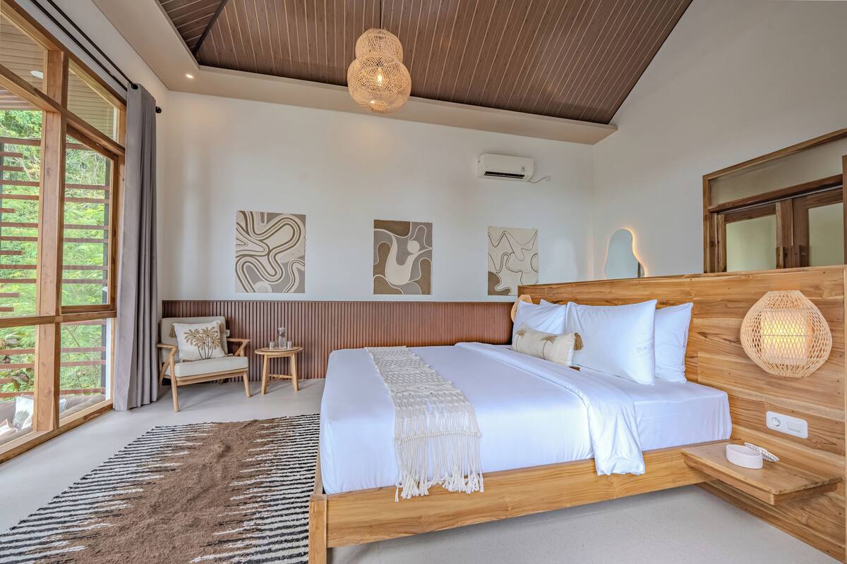 Quarto com uma cama grande, cabeceira de madeira e vista para o verde através de amplas janelas.