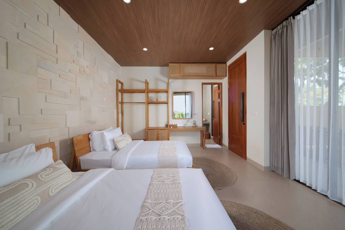 Quarto de hotel moderno com duas camas, teto de madeira, secretária embutida e uma janela grande com cortinas.