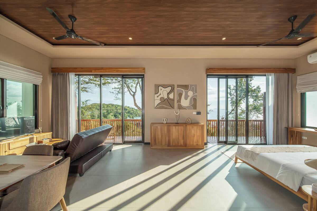 Quarto amplo com cama grande, sofá, mesa de jantar e televisão. Possui janelas grandes com cortinas e varanda com guarda-corpo de madeira.