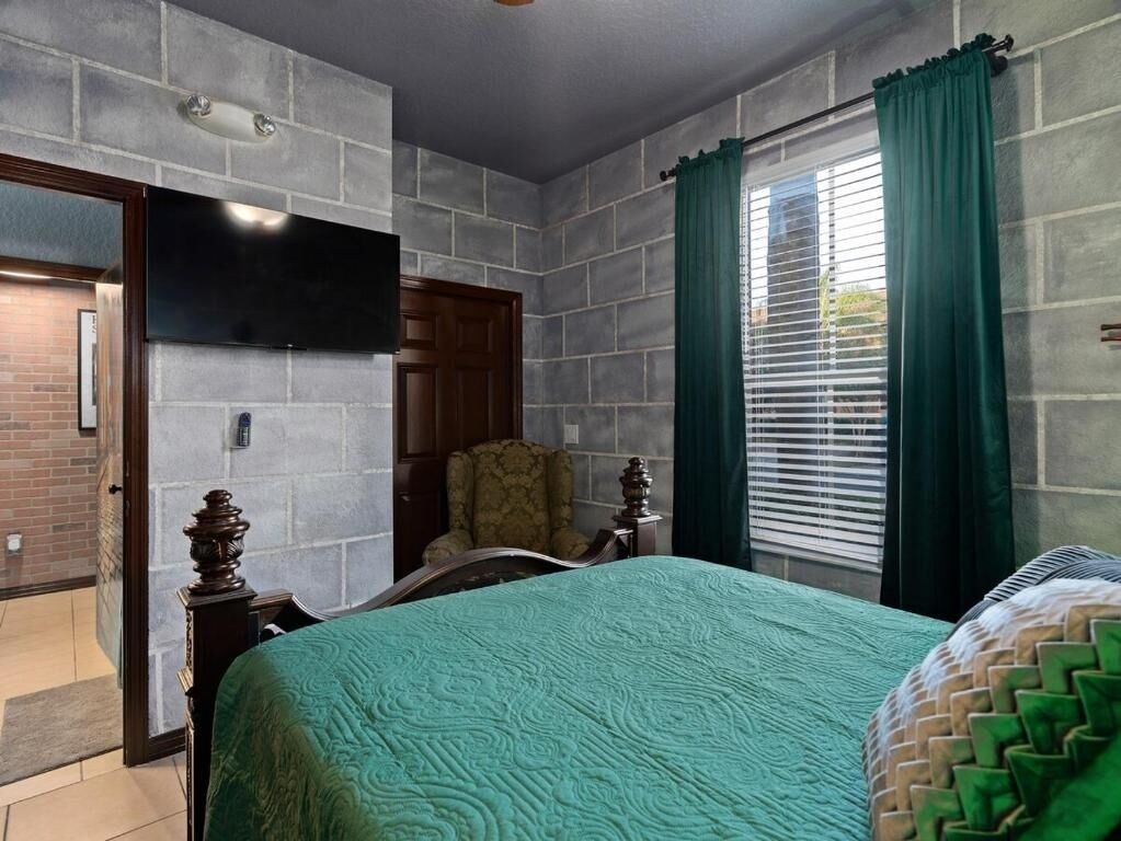Um quarto com uma cama, televisão na parede, uma cadeira e uma janela com cortinas.