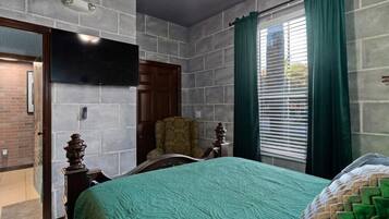 Um quarto com uma cama, televisão na parede, uma cadeira e uma janela com cortinas.