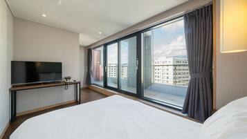 Quarto de hotel com uma cama grande, televisão, escrivaninha com telefone e vista para edifícios através de grandes janelas.