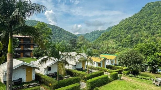 Paisagem verdejante e exuberante com várias tendas brancas e alguns edifícios ao fundo. Há sebes bem aparadas e uma pequena mesa com cadeiras em primeiro plano.