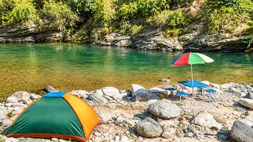 Um camping com tendas e uma mesa com cadeiras à beira de um rio calmo.