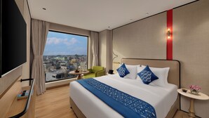 Quarto de hotel com uma cama grande, uma escrivaninha, uma cadeira, uma mesa pequena e uma janela ampla com vista para a cidade.