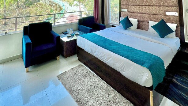 Quarto de hotel moderno com uma cama grande, ventilador de teto, área de estar com uma poltrona, e vista para árvores e uma ponte lá fora.