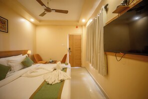 Quarto de hotel com cama, televisão, ventoinha no teto e porta que dá para outro cômodo.
