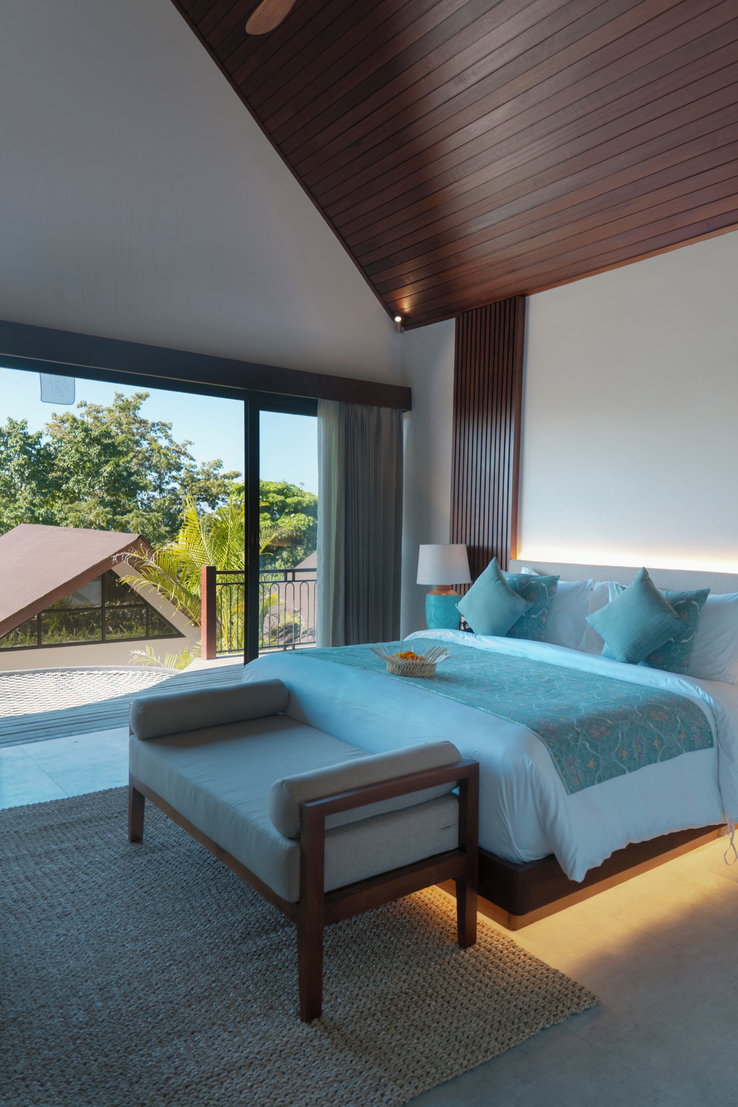 Quarto moderno com uma cama grande, um banquinho, uma mesinha de cabeceira e vista para o exterior.