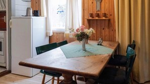 Uma mesa de jantar de madeira com cadeiras, um vaso com flores, um relógio na parede e uma área de cozinha com frigorífico e chaleira.
