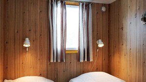 Quarto com paredes de madeira, duas camas com roupa de cama branca, janela com cortinas listradas e duas luzes fixadas na parede.