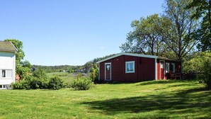 Uma cabana vermelha com porta e janelas, rodeada por um campo gramado e árvores, com um prédio branco ao fundo.