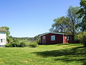 Uma cabana vermelha com porta e janelas, rodeada por um campo gramado e árvores, com um prédio branco ao fundo.