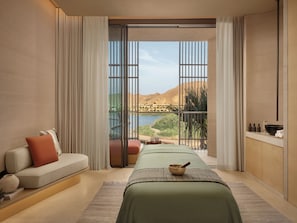 Quarto com cama, mesa de massagem, sofá, vista para um corpo de água e varanda com guarda-corrimão.