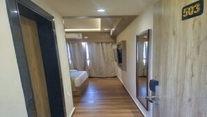 Corredor de hotel com piso de madeira, uma cama ao fundo e uma porta com placa de número.
