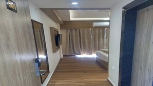 Um corredor com assoalho de madeira, porta com placa de identificação, televisão fixada na parede e janela com cortinas.