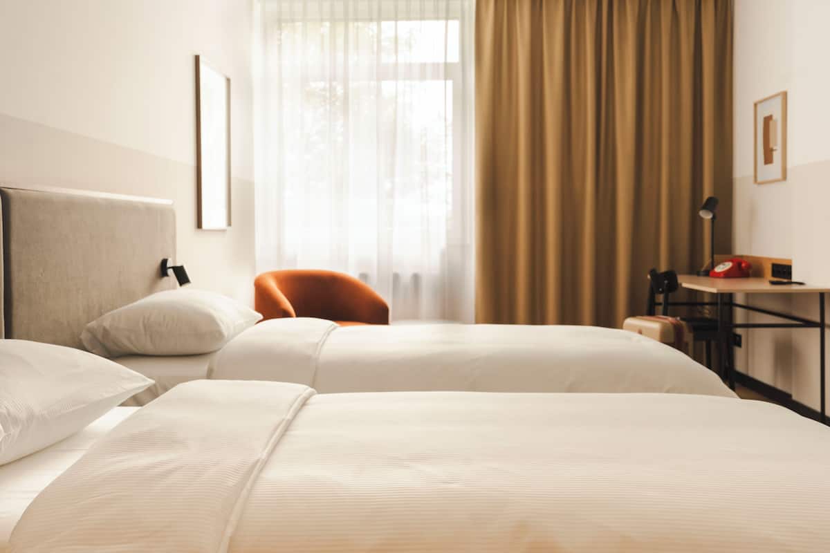 Quarto de hotel com duas camas, uma cadeira, uma escrivaninha e uma janela com cortinas.