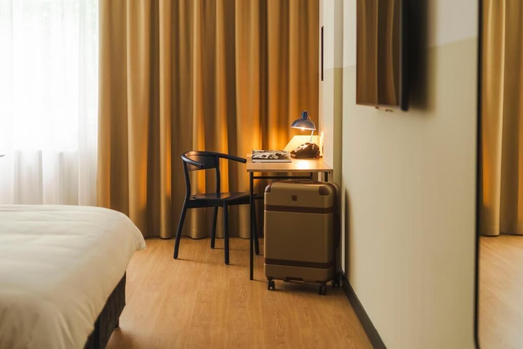 Quarto de hotel com uma cama, uma escrivaninha com luminária, uma cadeira e uma mala.