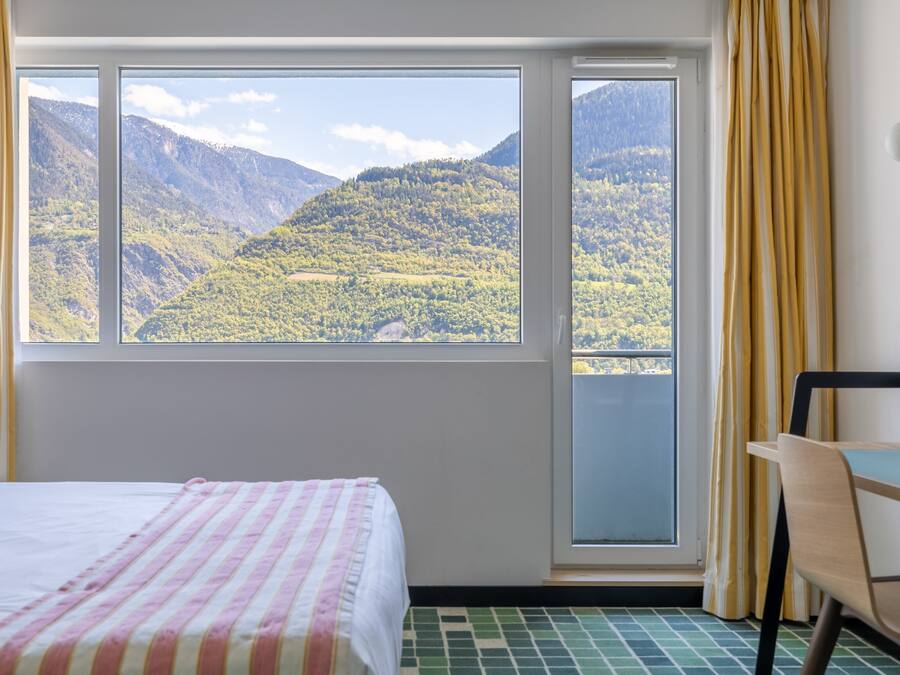 Quarto de hotel com cama, uma cadeira e uma grande janela com vista para uma paisagem montanhosa.