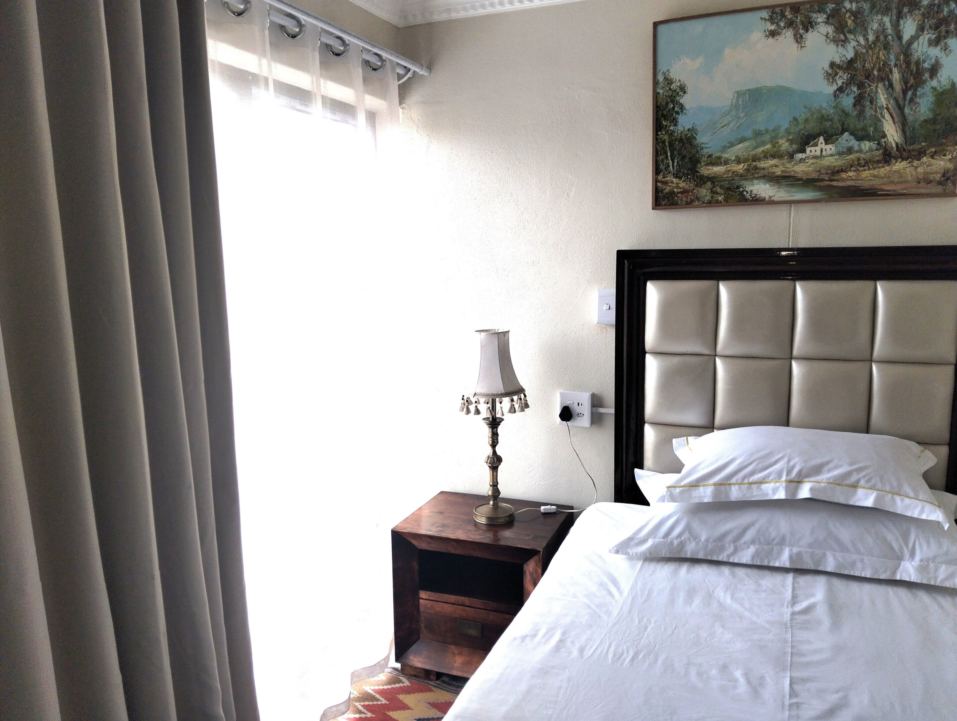 Quarto de hotel com cama, mesa de cabeceira, luminária, um quadro na parede e janela com cortinas.