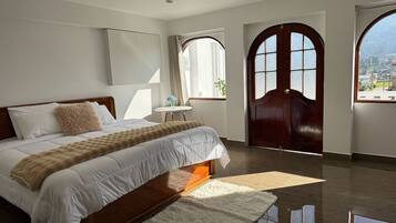 Quarto com uma cama grande, cabeceira de madeira, tapete branco e vista da cidade através de uma janela em arco.