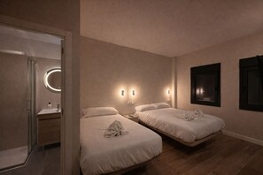 Quarto de hotel com duas camas, um espelho redondo, um banheiro pequeno e luminárias na parede.