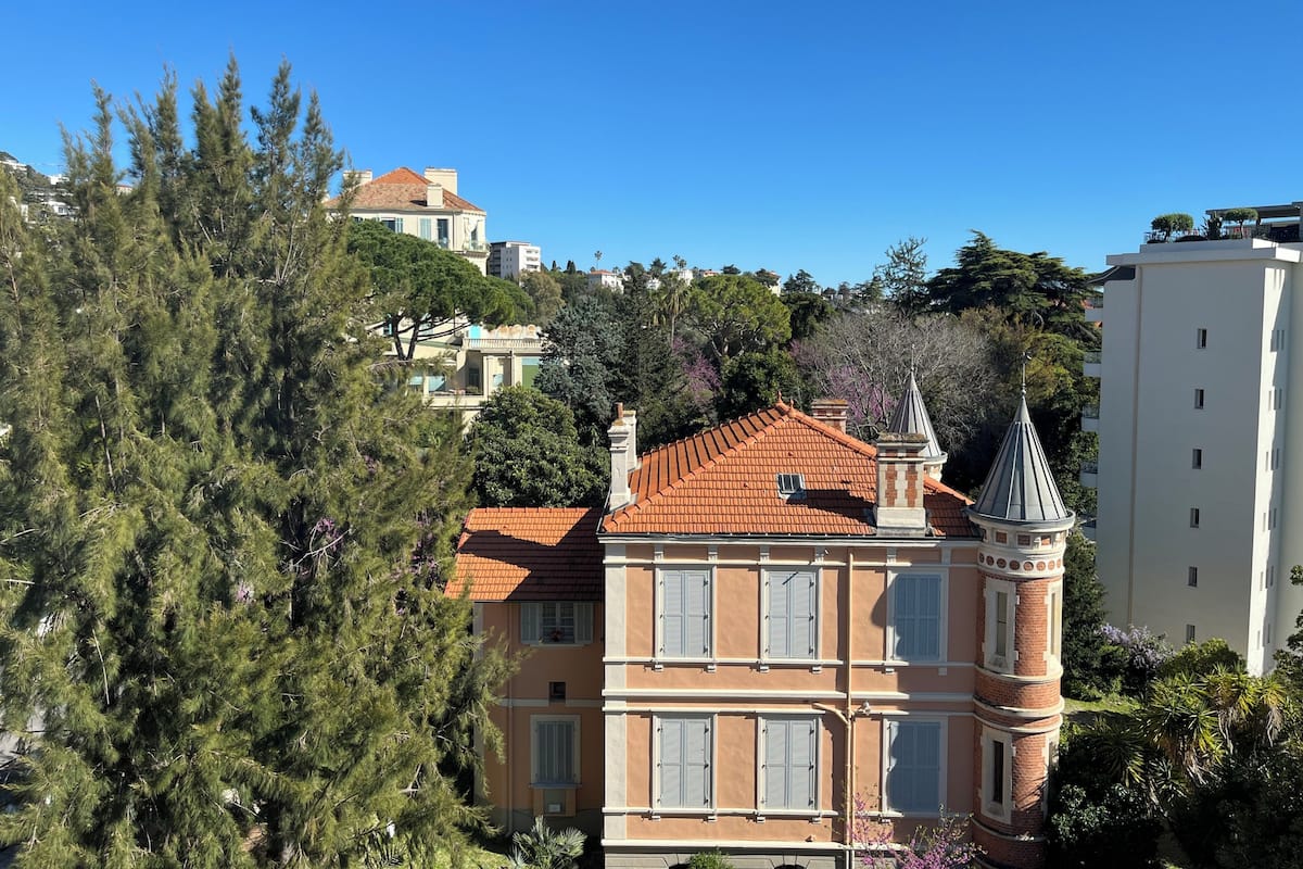 Um edifício com telhado de telhas vermelhas, uma torre com telhado cónico e uma grande árvore em primeiro plano.