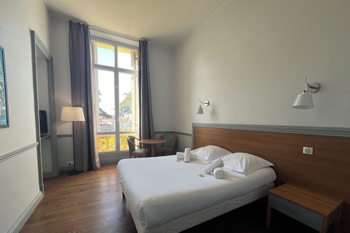 Quarto de hotel com uma cama grande, mesas de cabeceira, uma cadeira, janela com cortinas e vista para o exterior.