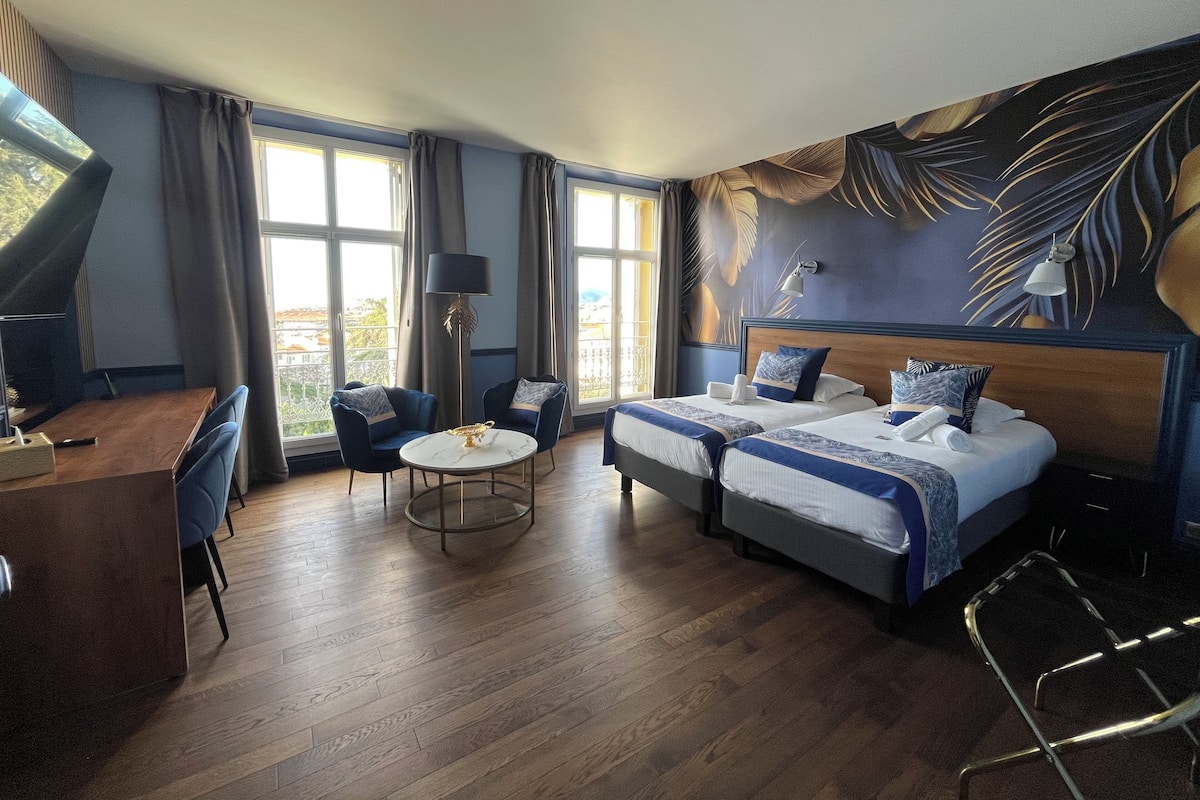 Quarto de hotel com duas camas, uma escrivaninha, uma cadeira, uma mesinha e janelas grandes com cortinas.