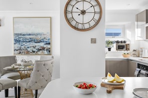 Cozinha moderna com um relógio grande na parede, área de jantar com mesa redonda e cadeiras, e vista para o mar pela janela.