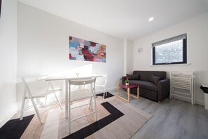 Um espaço de estar moderno com uma mesa branca, cadeiras, um sofá cinzento, um quadro, uma janela e um radiador.