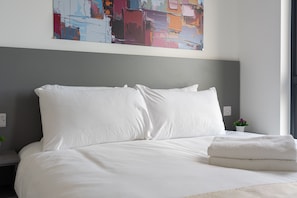 Quarto de hotel com cama grande, cabeceira cinza e uma pintura abstrata colorida acima da cama.