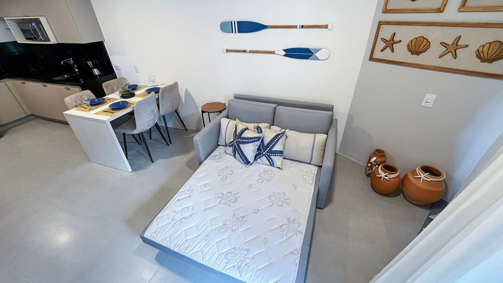 Quarto pequeno com cama, escrivaninha e cadeiras. Há objetos decorativos na parede e dois vasos no chão.