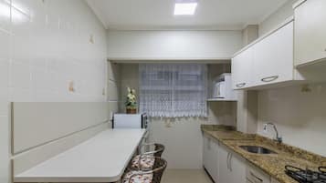 Cozinha compacta com armários brancos, bancada de granito e uma pequena janela com cortinas de renda.