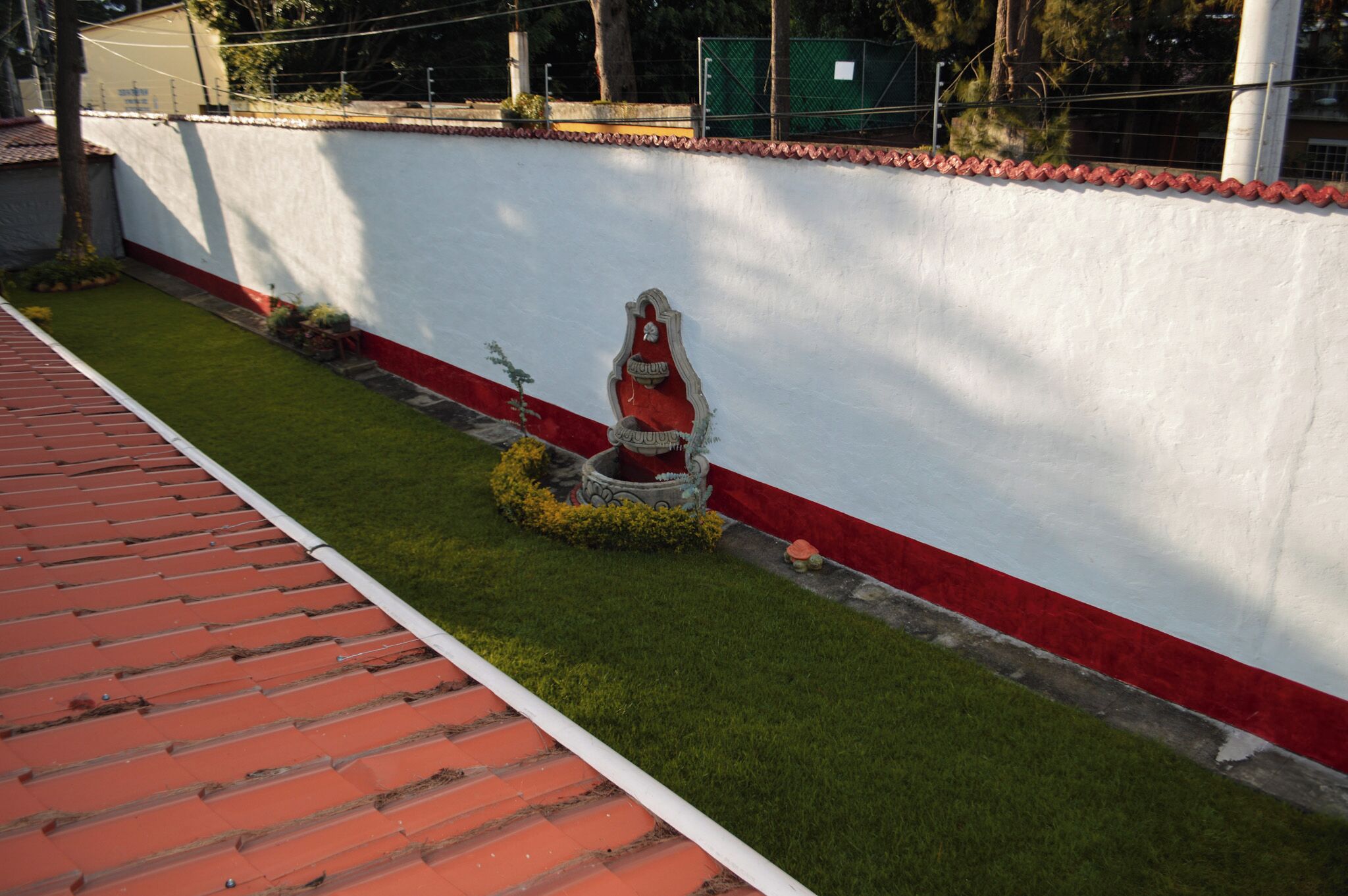 Terraço com telhas vermelhas, parede branca com detalhes vermelhos e um pequeno jardim com um vaso decorativo.