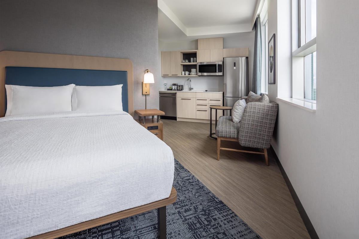 Quarto de hotel com cama, uma mesinha, uma cadeira, cozinha compacta e janela com cortinas.