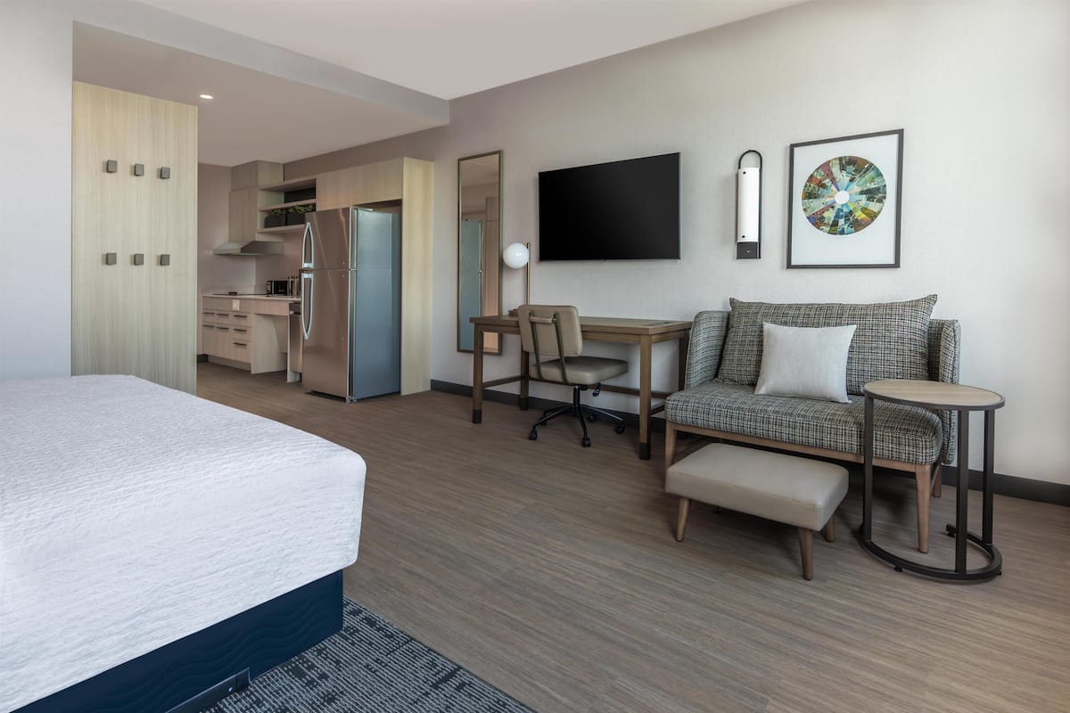 Quarto de hotel moderno com cama, sofá, escrivaninha, cadeira, TV e cozinha.