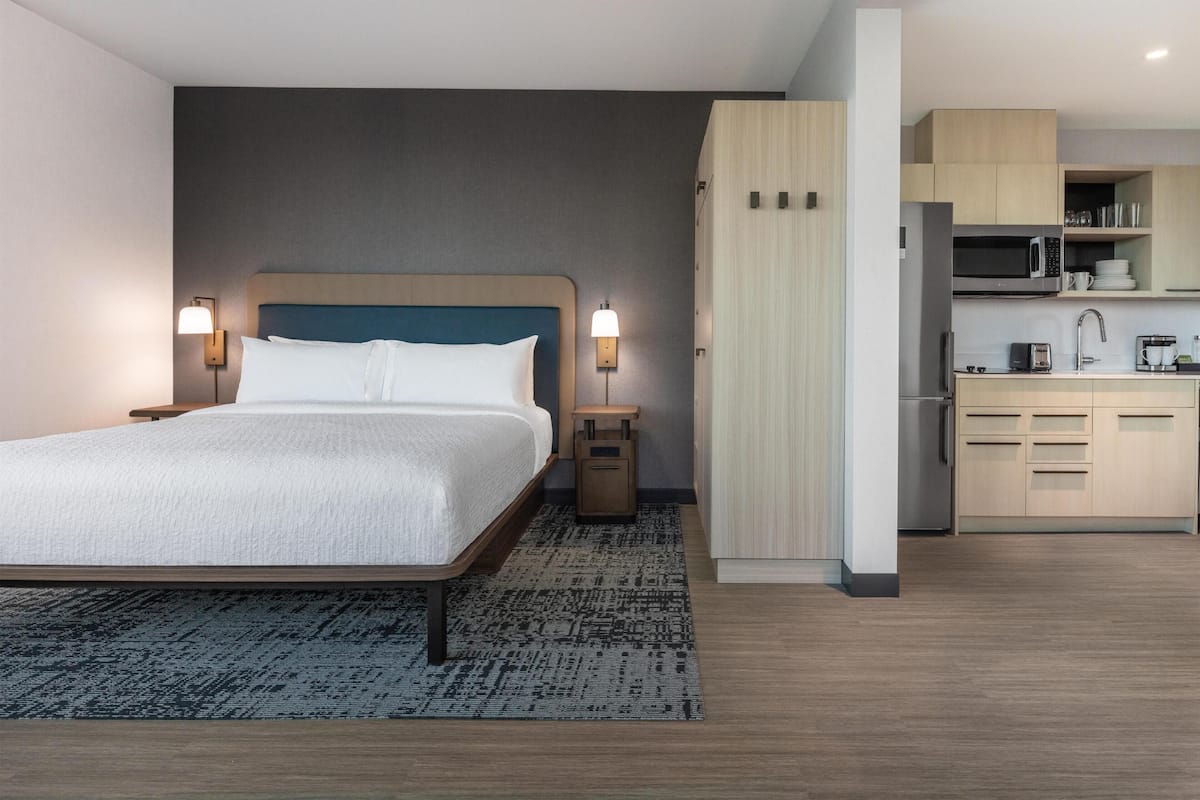 Quarto de hotel com cama, abajures, mesa de cabeceira pequena, área de cozinha com micro-ondas e armário.