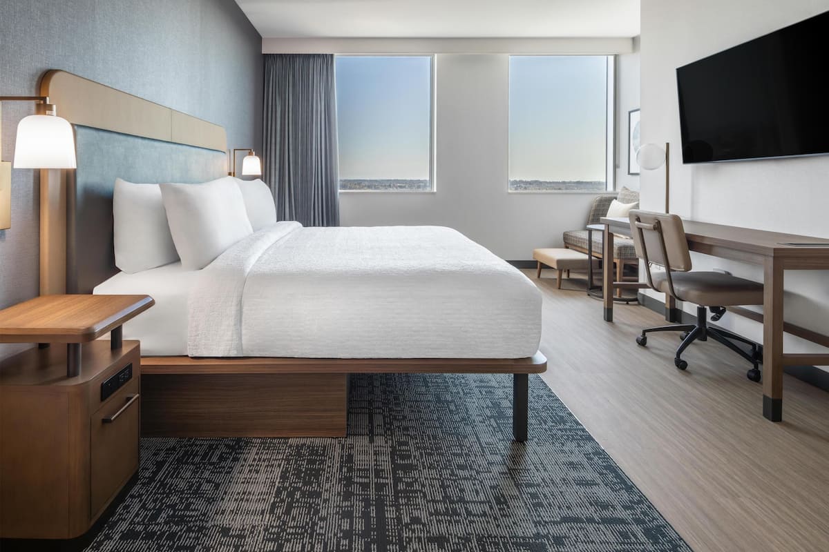 Quarto de hotel com cama, escrivaninha, cadeira, televisão e janelas amplas.