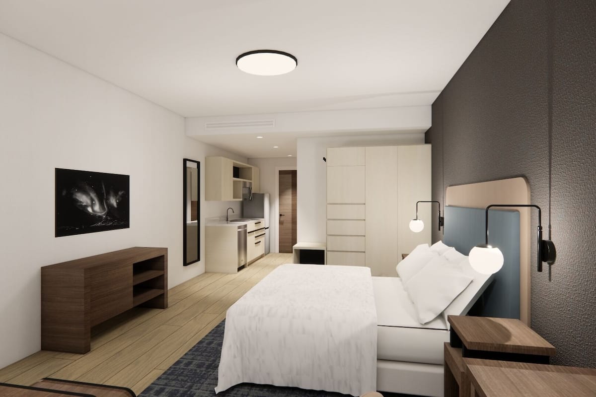 Quarto de hotel moderno com uma cama grande, cômoda de madeira, escrivaninha e cozinha compacta.