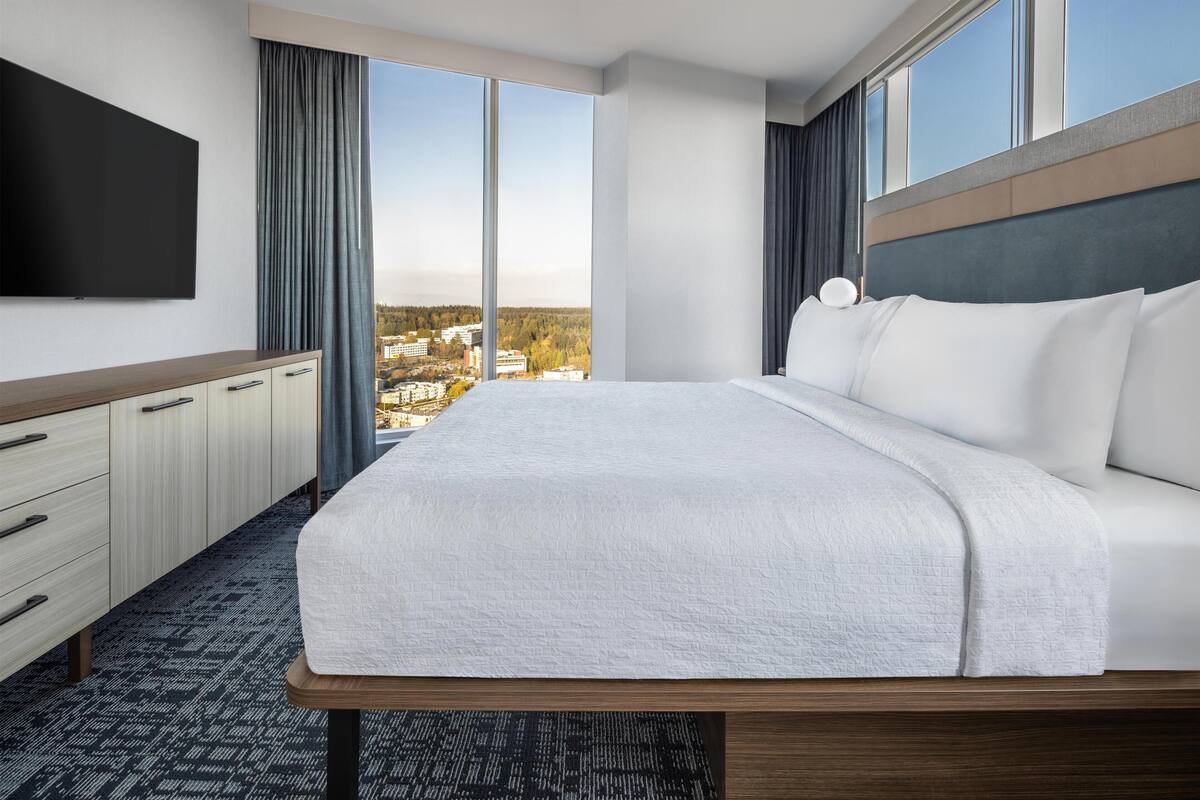 Quarto de hotel com uma cama grande, televisão de tela plana, cômoda de madeira e vista para a paisagem urbana através de grandes janelas.